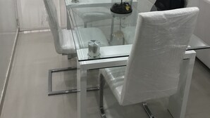 Uma área de refeições moderna com mesa de vidro, cadeiras de metal e um vaso de vidro com uma flor amarela.