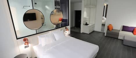 Quarto de hotel moderno com uma cama grande, um espelho redondo, uma pequena mesa e um sofá com almofadas coloridas.