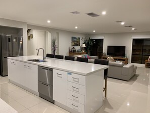 Cozinha moderna com ilha central, máquina de lavar louça embutida e área de jantar com sofá e televisão.