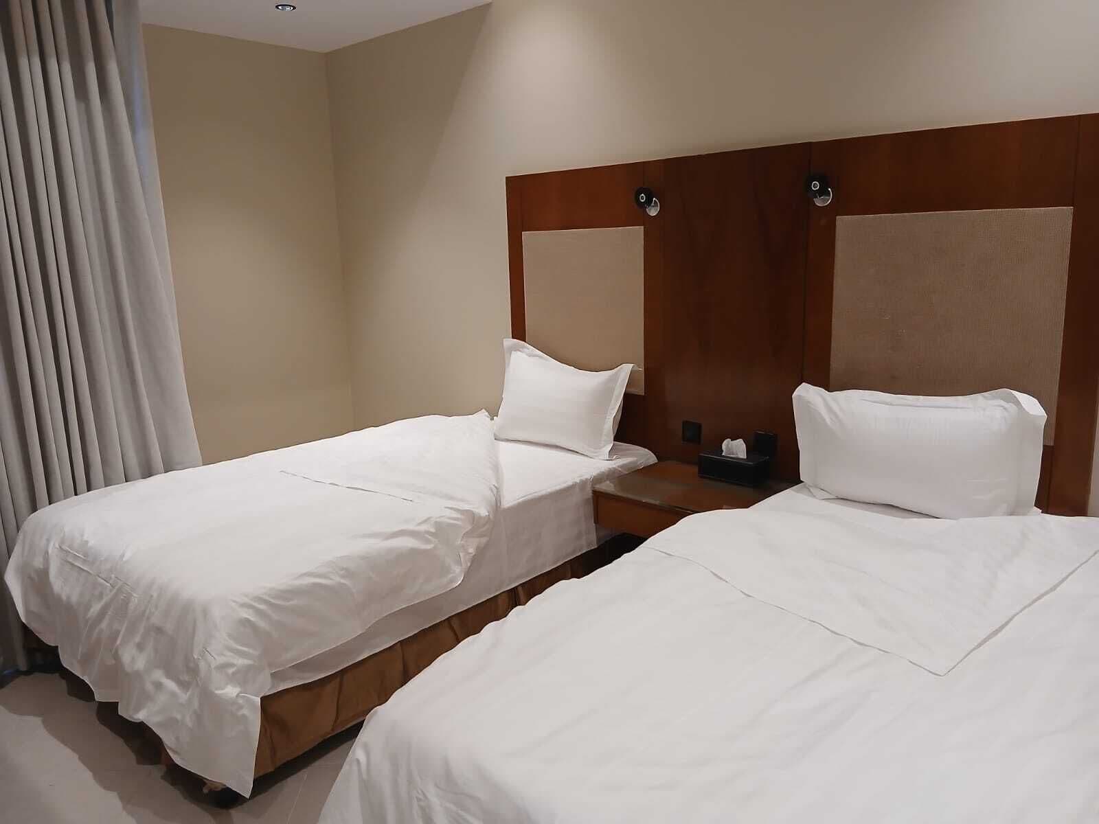 Quarto de hotel com duas camas de solteiro, cabeceira de madeira e mesa de cabeceira com luminária.