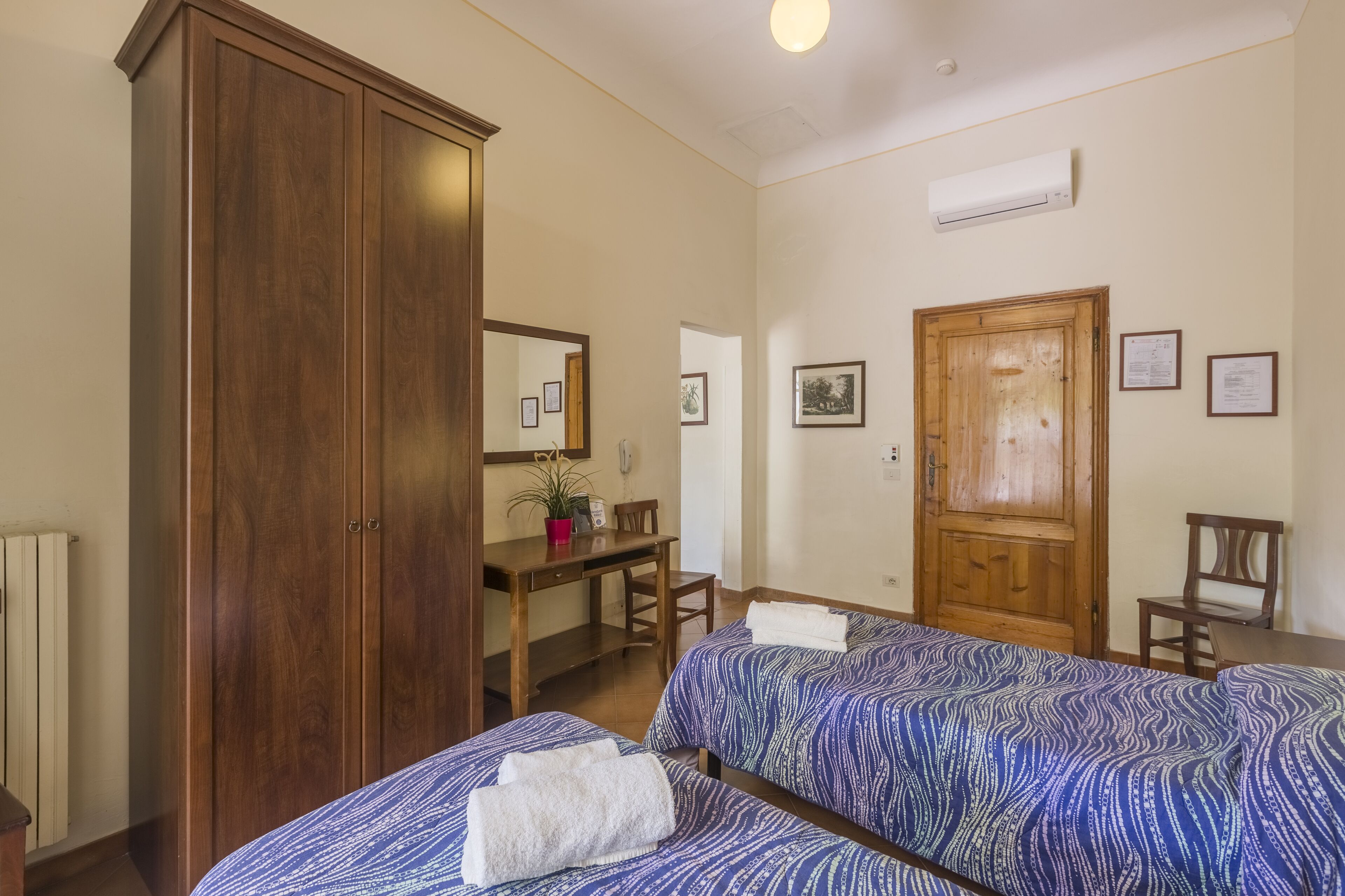 Quarto com duas camas, um guarda-roupa de madeira, uma escrivaninha com cadeira, um vaso de planta e uma porta de madeira.