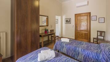 Quarto com duas camas, um guarda-roupa de madeira, uma escrivaninha com cadeira, um vaso de planta e uma porta de madeira.