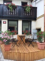 Um deck de madeira ao ar livre com mesa e cadeiras, rodeado por vasos com plantas e flores.
