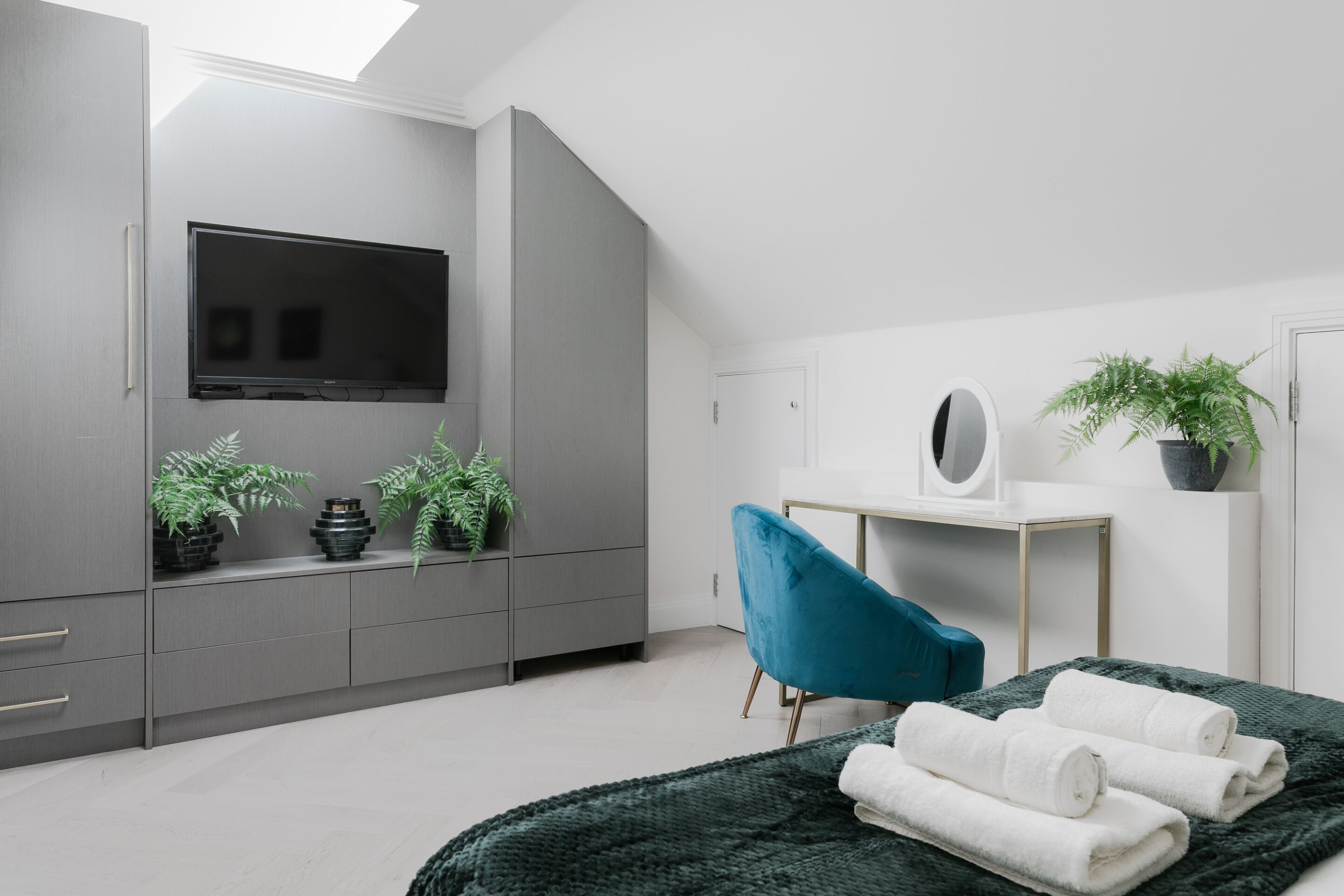 Um quarto moderno com um guarda-roupa cinzento, uma televisão de ecrã plano, uma cadeira azul, uma cama com toalhas brancas dobradas e uma planta num vaso.