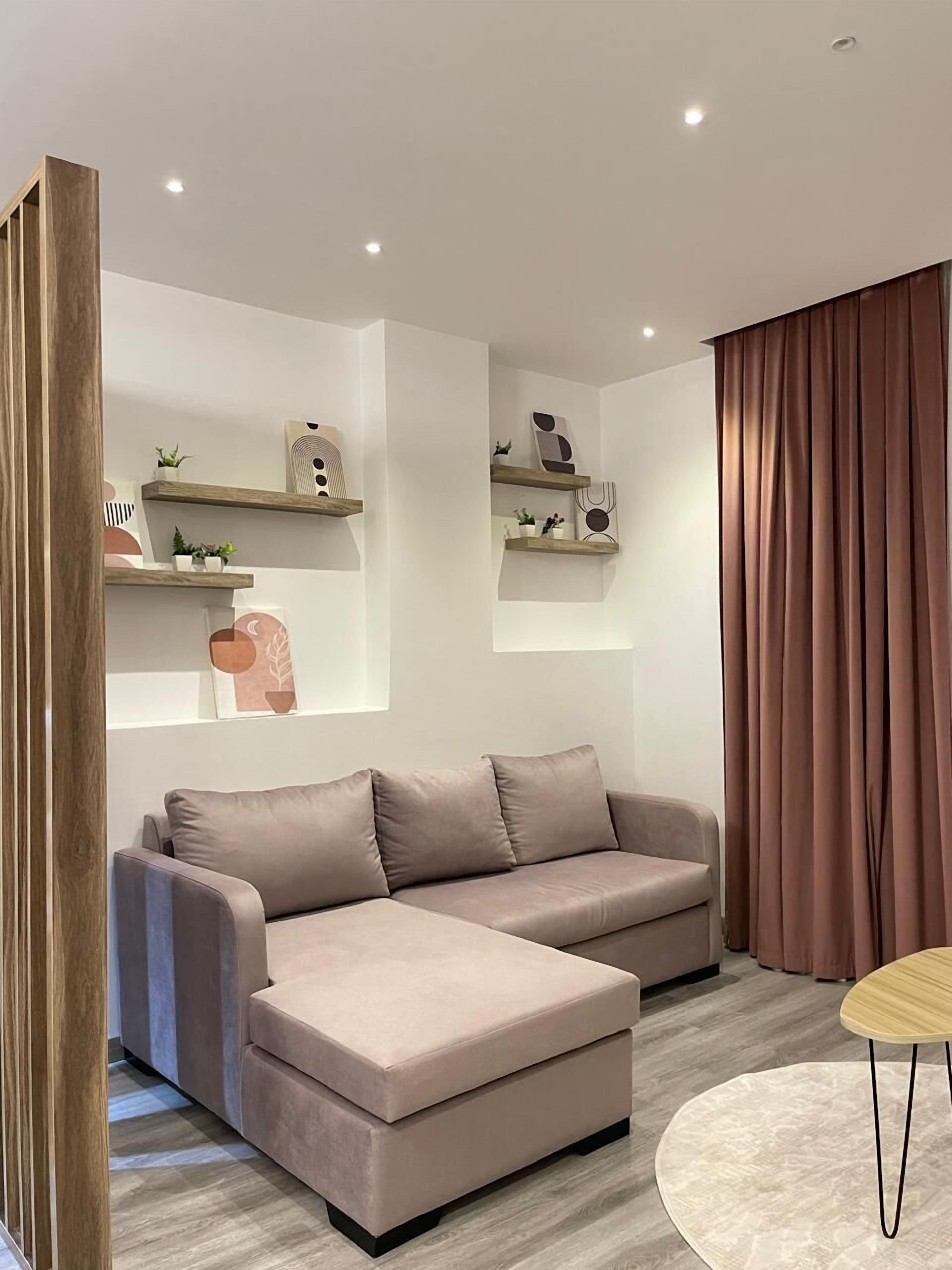 Uma sala de estar moderna com um sofá seccional bege, prateleiras de madeira com objetos decorativos e uma mesa de centro redonda de madeira.