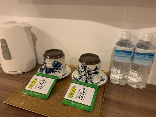 Uma área de cozinha com uma chaleira elétrica branca, duas xícaras de cerâmica com desenhos florais azuis, duas garrafas de água e dois sachês de chá sobre um suporte.