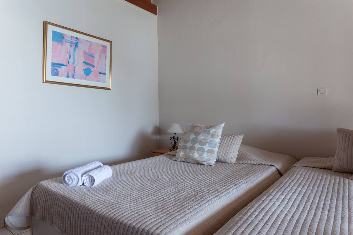 Quarto de hotel com duas camas, um quadro na parede e uma mesa de cabeceira com abajur.