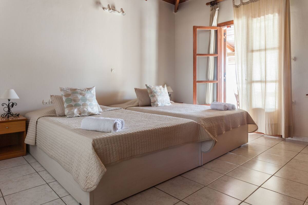 Quarto com duas camas, mesa de cabeceira, abajur e janela com cortinas.