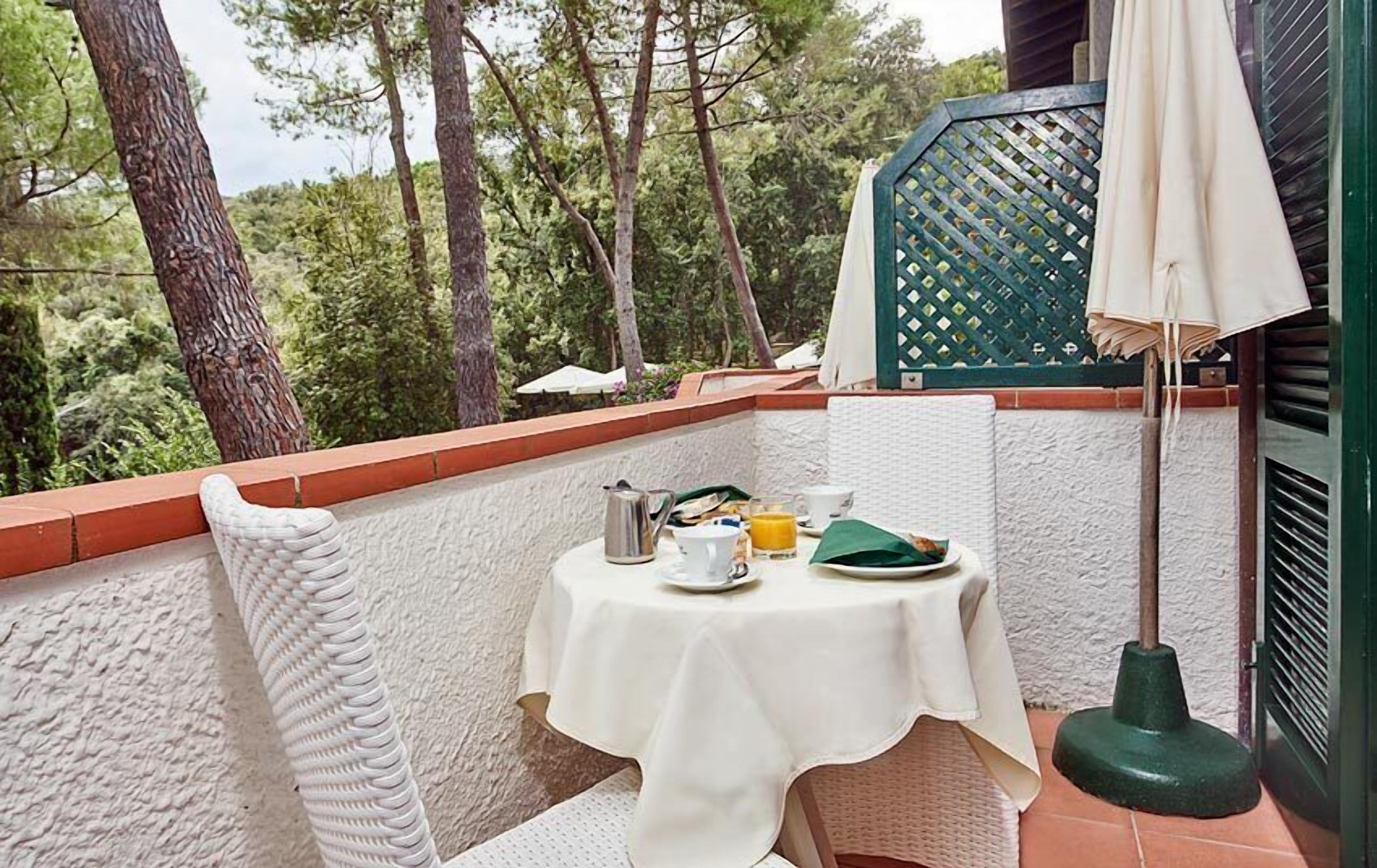 Uma varanda com mesa posta para o café da manhã, um guarda-sol verde e vista para árvores.
