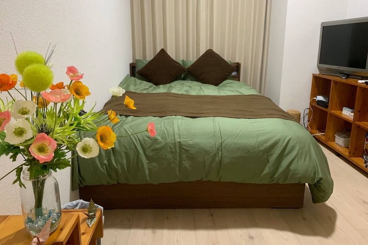 Quarto bem arrumado com colcha verde, duas almofadas marrons, mesa de cabeceira de madeira com um vaso de flores e uma televisão em uma estante de madeira.
