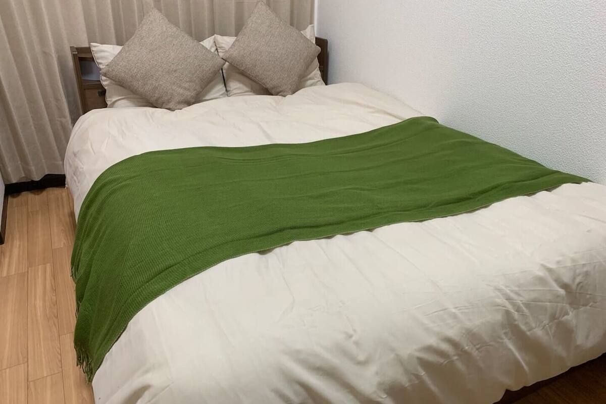 Cama bem arrumada com manta verde e duas almofadas, mesa de cabeceira de madeira com abajur, e janela com persianas.