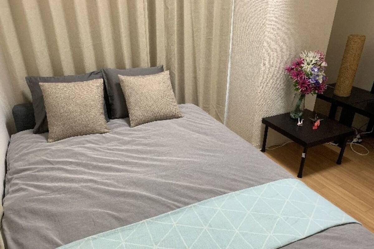 Cama bem arrumada com edredom cinza e almofadas, uma mesinha de cabeceira com um vaso de flores, e piso de madeira.