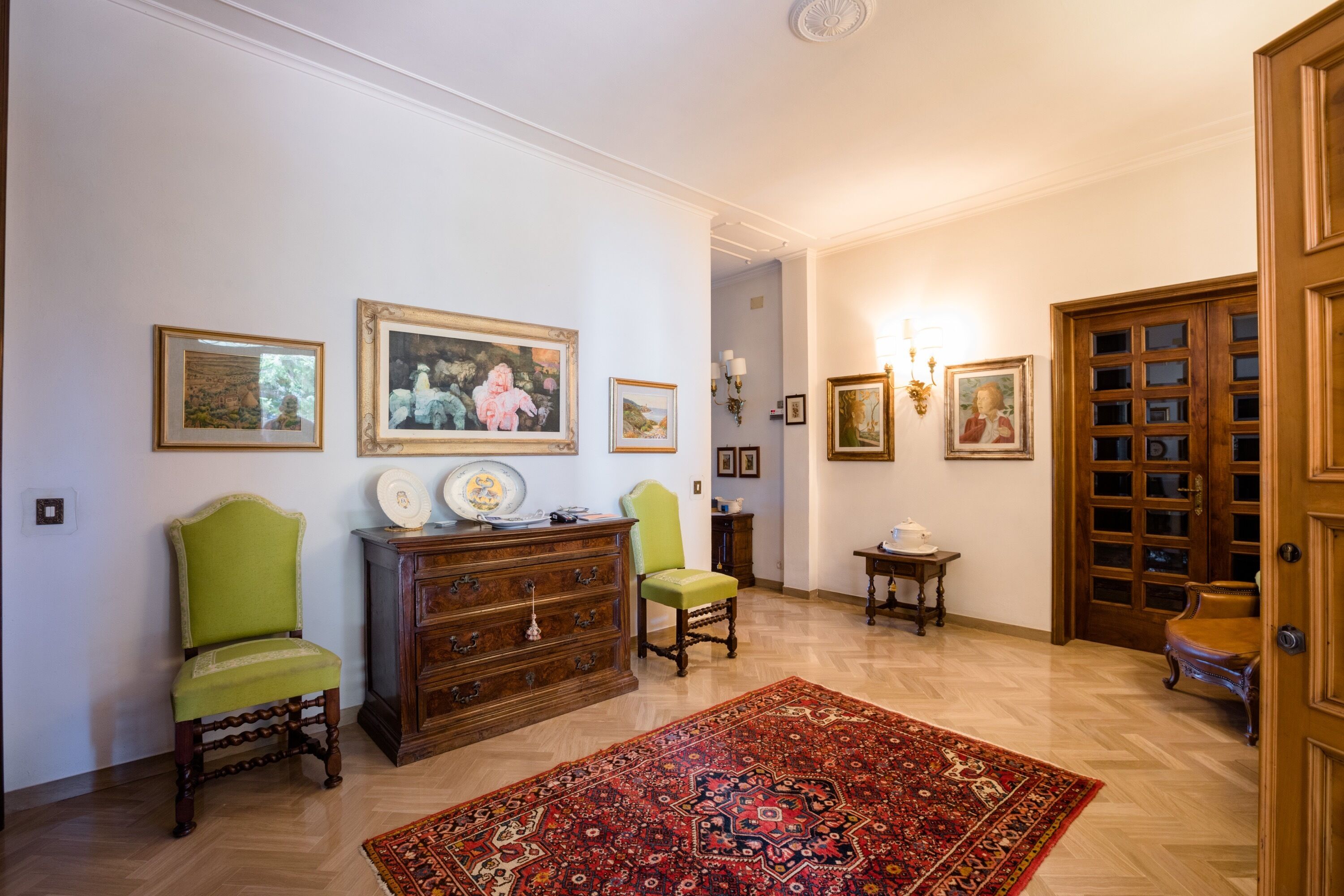 Um corredor com piso de madeira, cadeiras verdes, um tapete estampado em vermelho e quadros nas paredes.