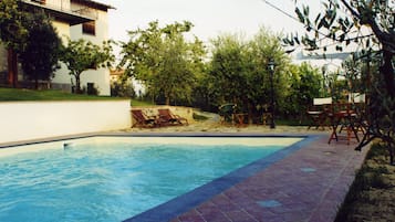 Piscina com deck ladrilhado, rodeada por árvores e mobiliário de exterior.