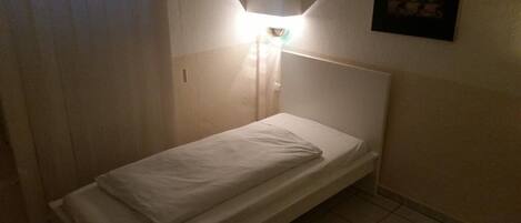 Quarto pequeno com uma cama de solteiro, cabeceira branca e luminária de cabeceira.
