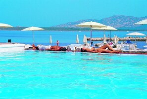 Área de piscina com espreguiçadeiras e guarda-sóis, piscina azul límpida e montanhas ao fundo.
