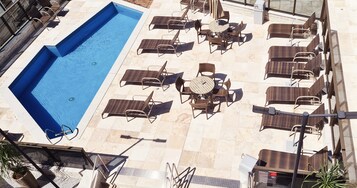 Vista aérea de uma área de piscina de hotel, com uma piscina retangular, várias espreguiçadeiras e mesas dispostas ao redor.