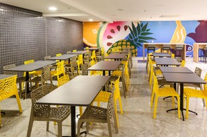 Uma área de refeições com várias mesas e cadeiras, parede revestida de azulejos e um mural colorido na parede de fundo.
