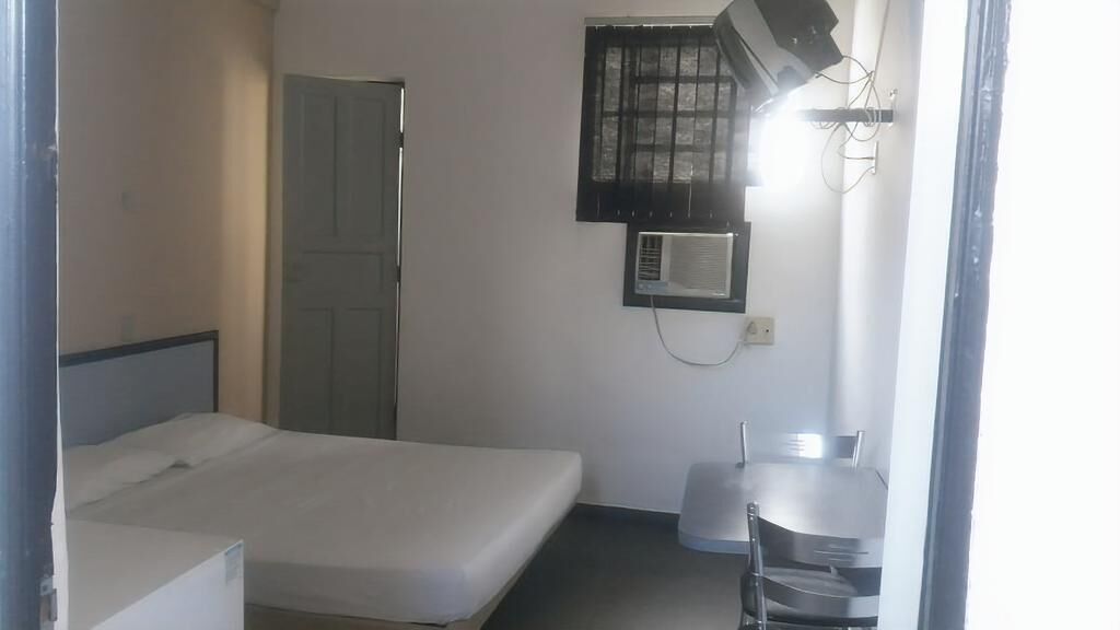 Um quarto pequeno e simples, com uma cama, uma escrivaninha, uma cadeira, uma porta e um ar condicionado.