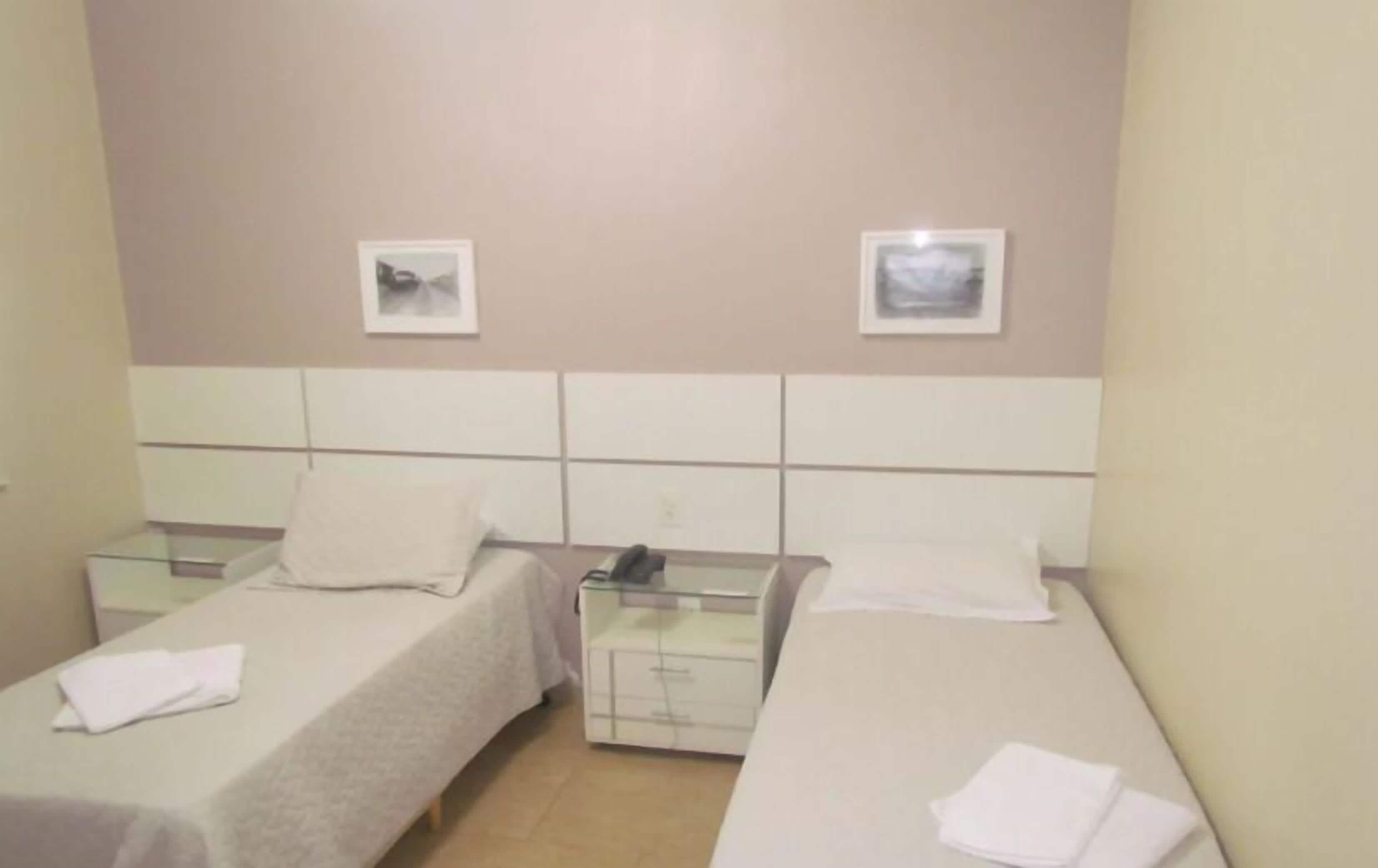 Quarto de hotel com duas camas de solteiro, uma mesa de cabeceira com abajur, e duas fotos emolduradas na parede.