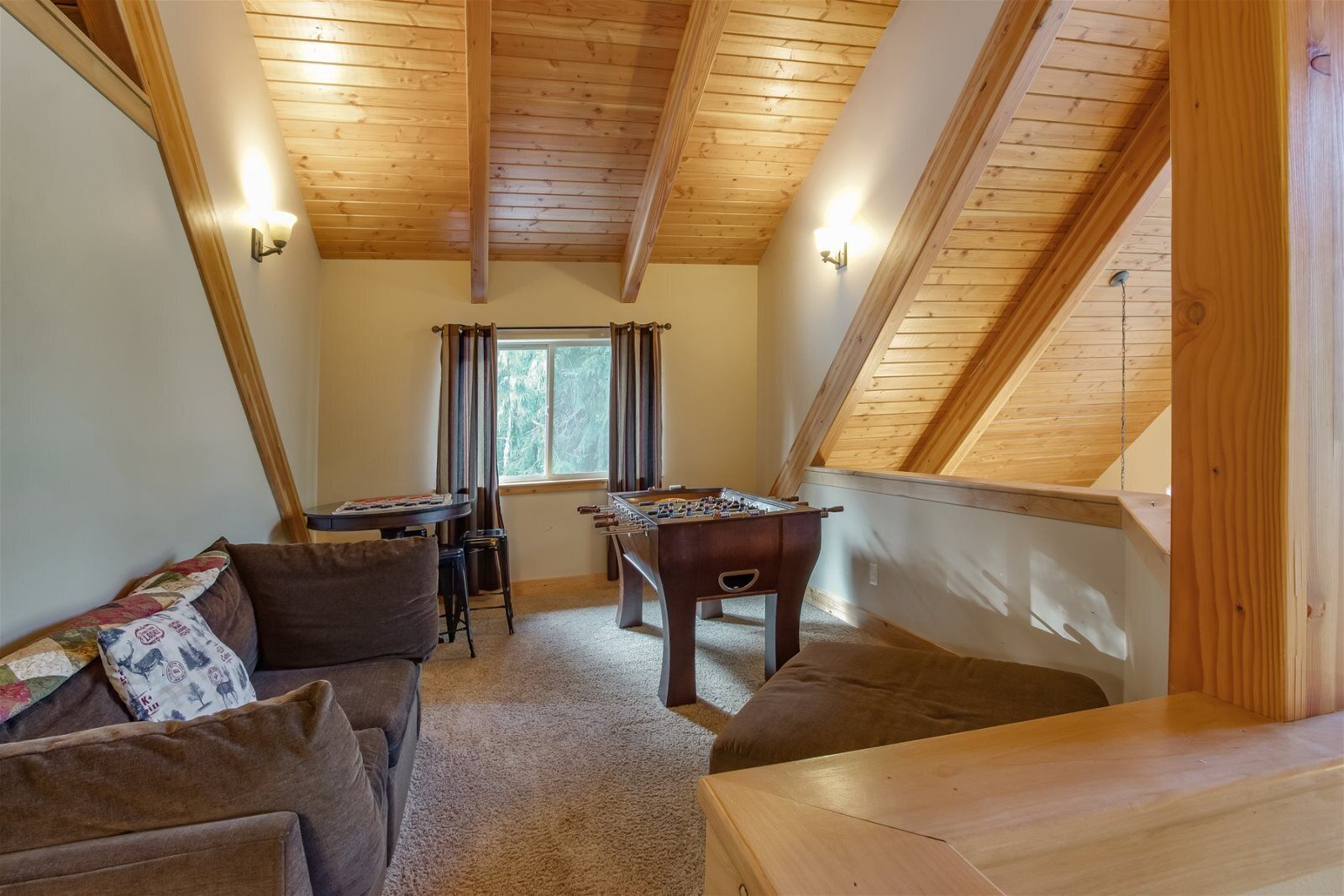 Quarto tipo loft acolhedor, com teto de madeira, mesa de futebol, pequena mesa de jantar e sofá.