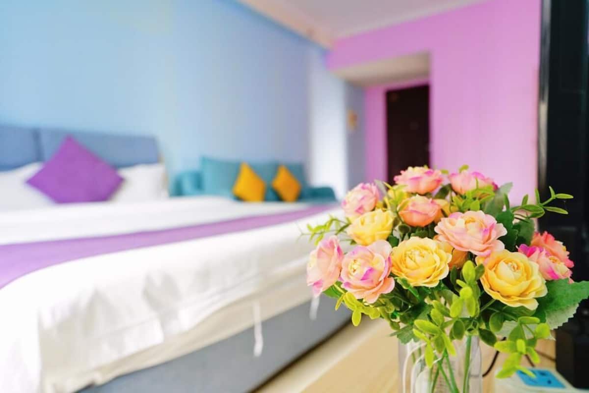 Uma cama com colcha e almofadas brancas, um vaso com flores rosa e amarelas, e um quarto com paredes azuis e rosa.