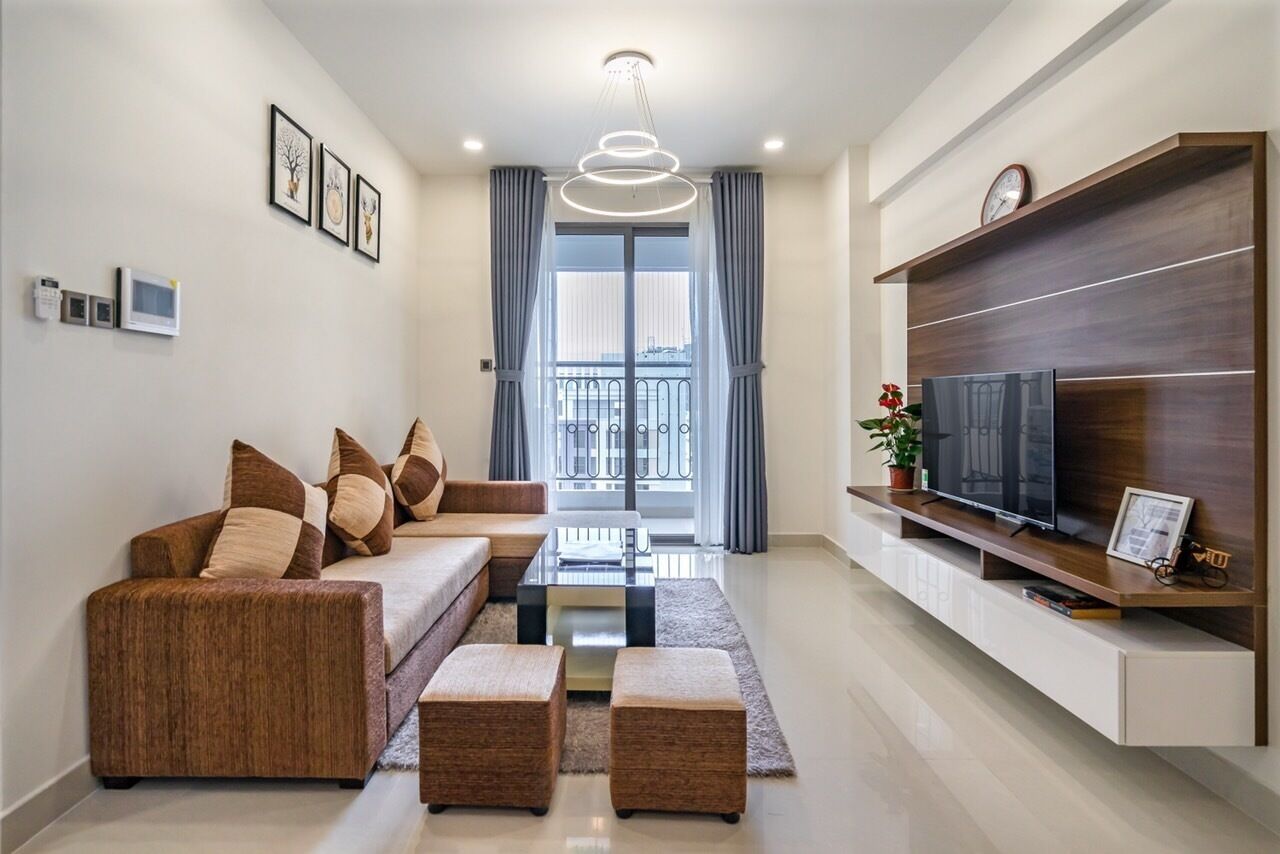 Uma sala de estar moderna com sofá, pufes, uma mesa de centro, uma TV em uma prateleira de madeira e uma varanda com cortinas.