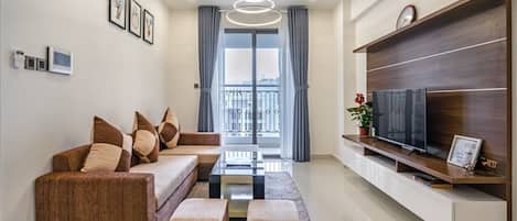 Uma sala de estar moderna com sofá, pufes, uma mesa de centro, uma TV em uma prateleira de madeira e uma varanda com cortinas.