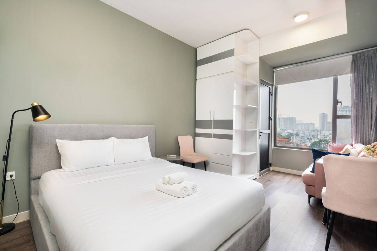 Quarto de hotel com cama, cadeira, escrivaninha e uma janela grande com vista para a cidade.