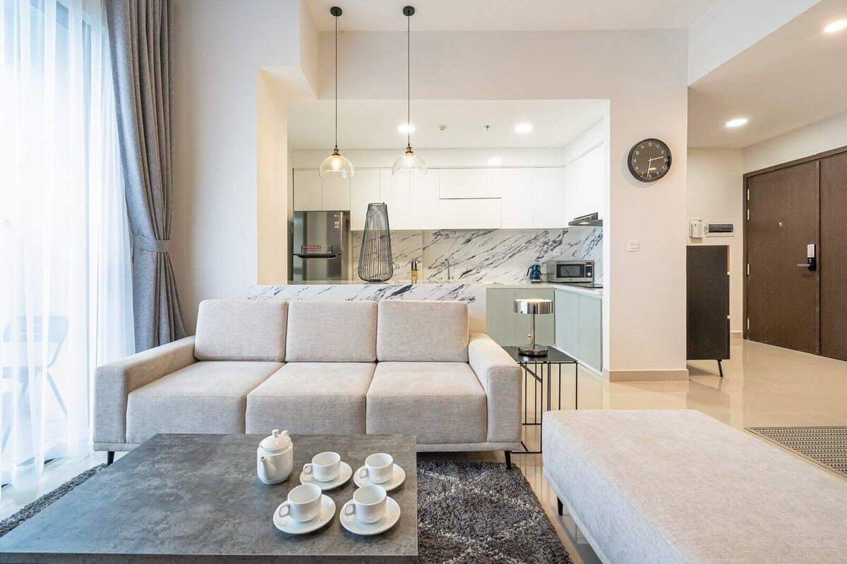Uma sala de estar moderna com um sofá, uma mesa de centro com chávenas e um bule, uma área de cozinha com um micro-ondas, e um relógio na parede.