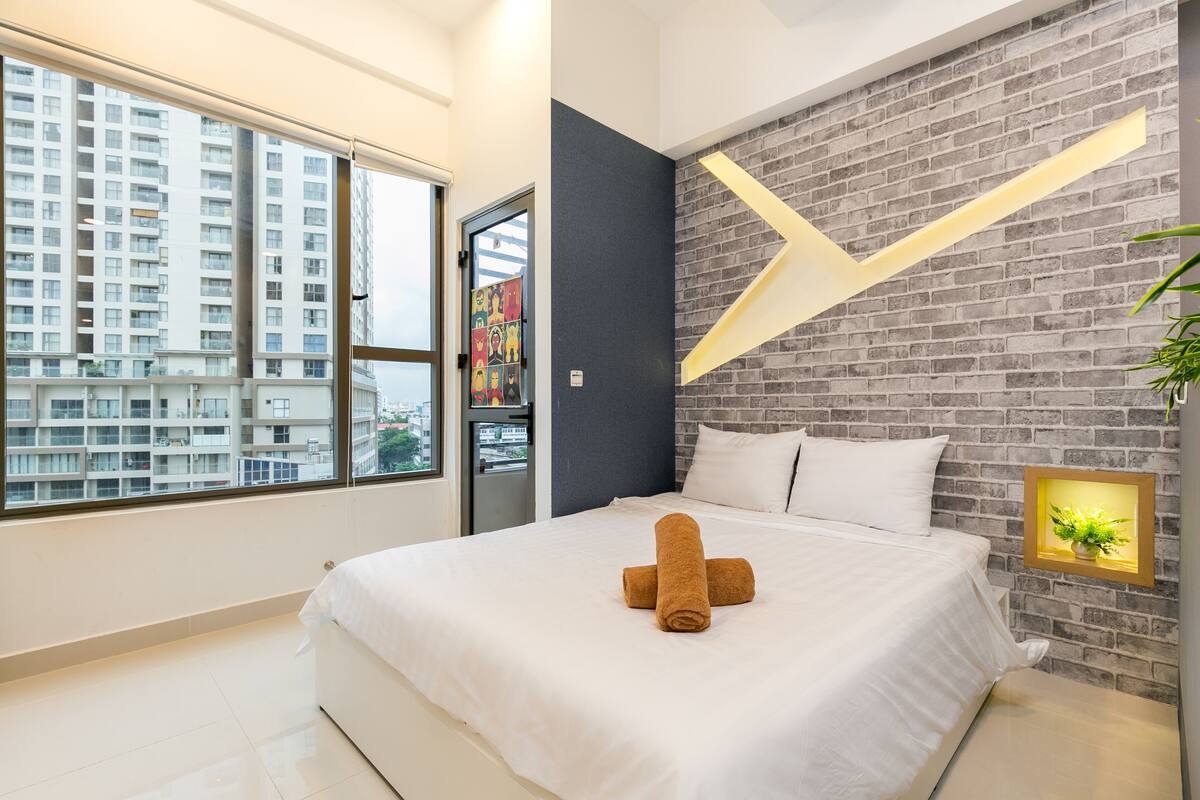 Quarto de hotel moderno com uma grande janela, parede de tijolos decorativa e uma cama bem arrumada com toalhas dobradas.