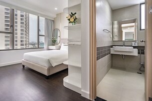 Quarto de hotel moderno com cama grande, chuveiro amplo, toucador com espelho e vista para a cidade através de grandes janelas.