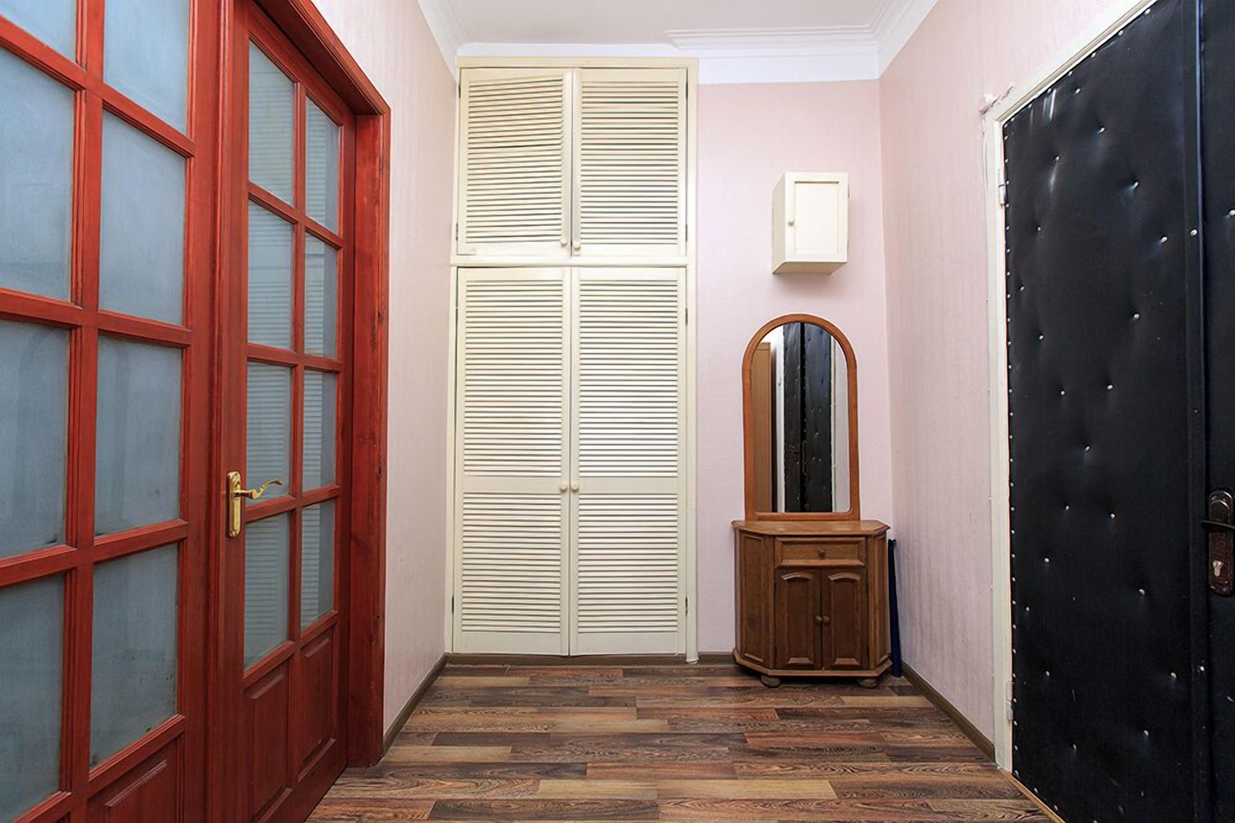 Um corredor com piso de madeira, uma porta vermelha, uma porta branca, uma porta preta e um espelho sobre um armário de madeira.