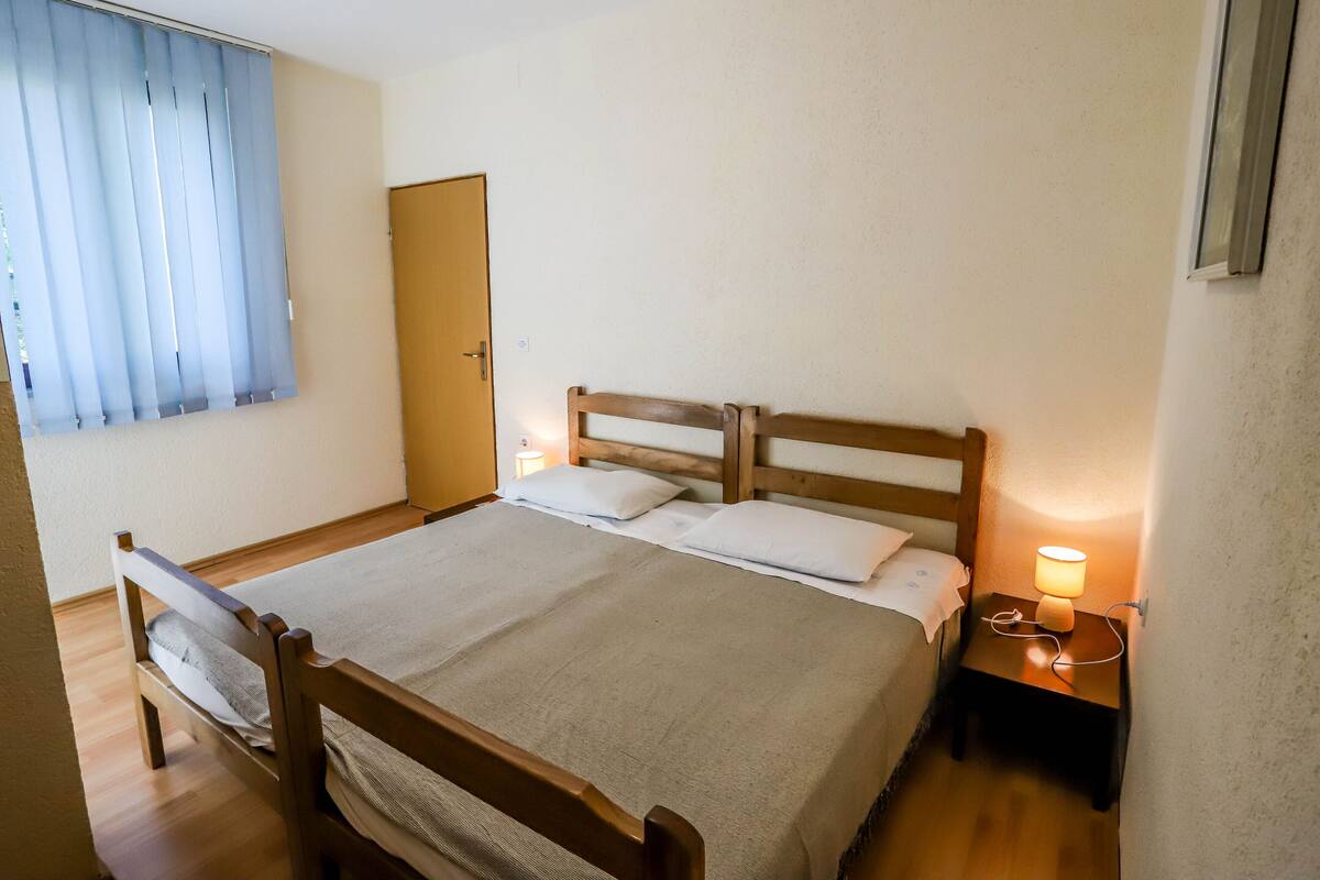 Cama bem arrumada com cabeceira de madeira, mesa de cabeceira com abajur e janela com persianas.