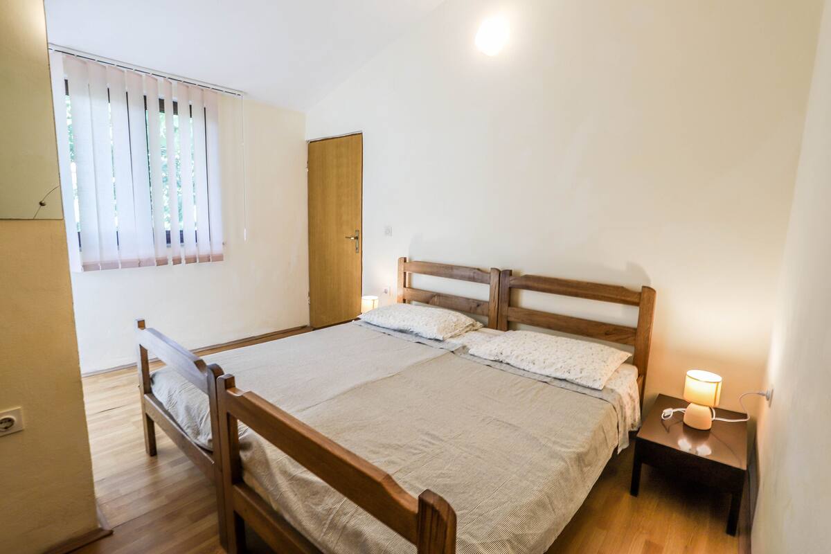 Quarto com cama de madeira, mesa de cabeceira com abajur e janela com persianas.