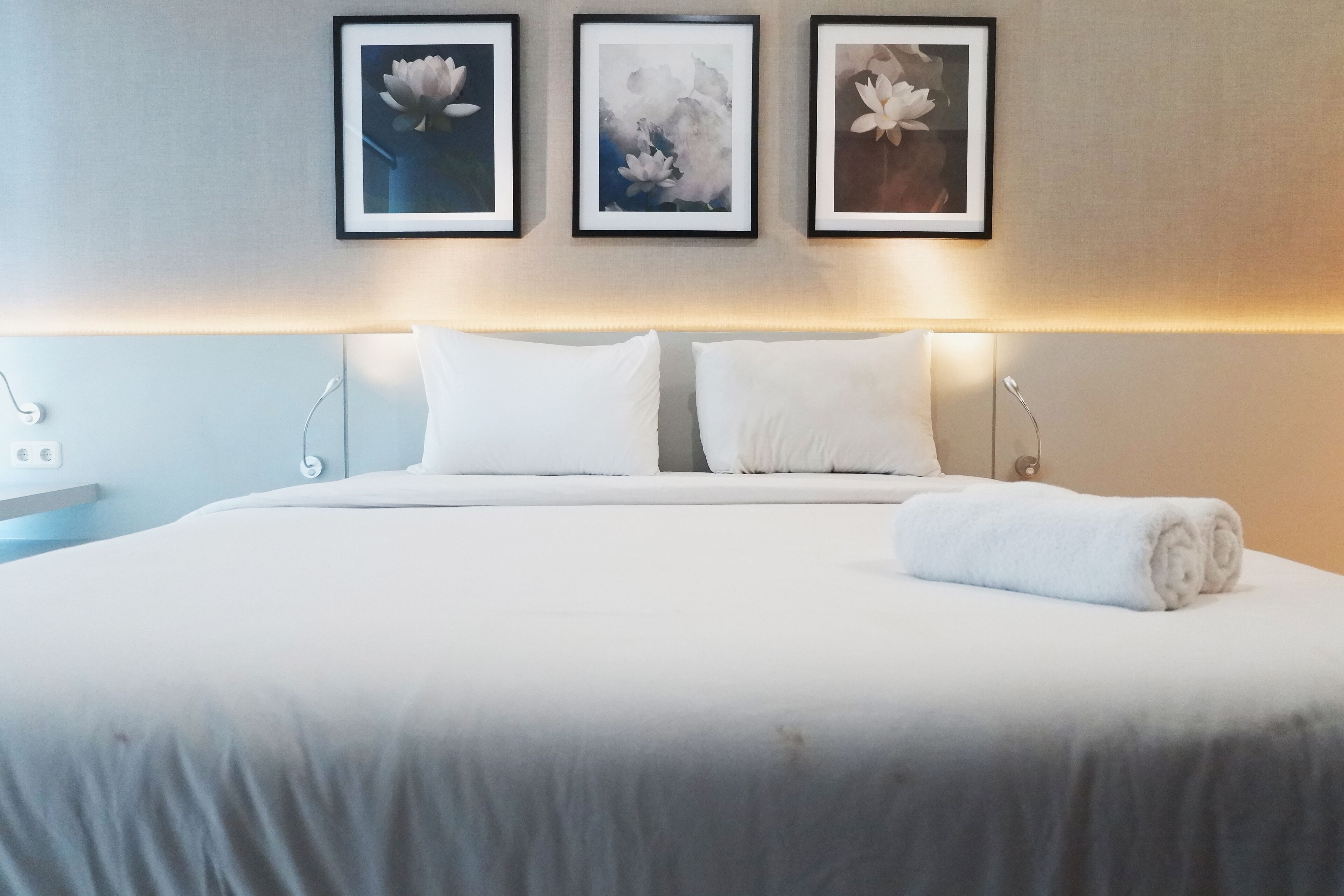 Uma cama bem arrumada com lençóis brancos e uma toalha enrolada, uma mesinha de cabeceira com abajur, e três quadros florais na parede.