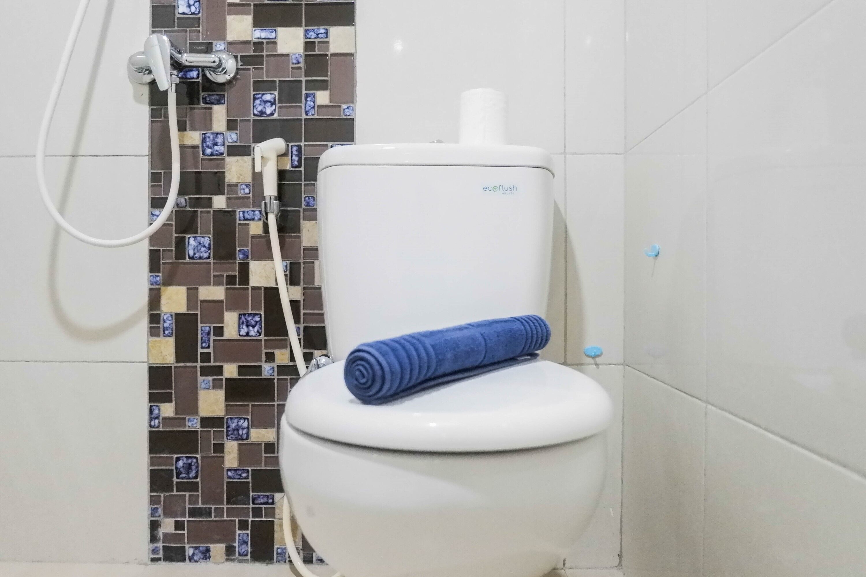 Casa de banho com toalha azul, mangueira de duche e parede de azulejos mosaicados.