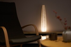 Uma cadeira com almofada escura, uma mesa com um vaso e uma vela, e um lustre alto e fino em uma prateleira.