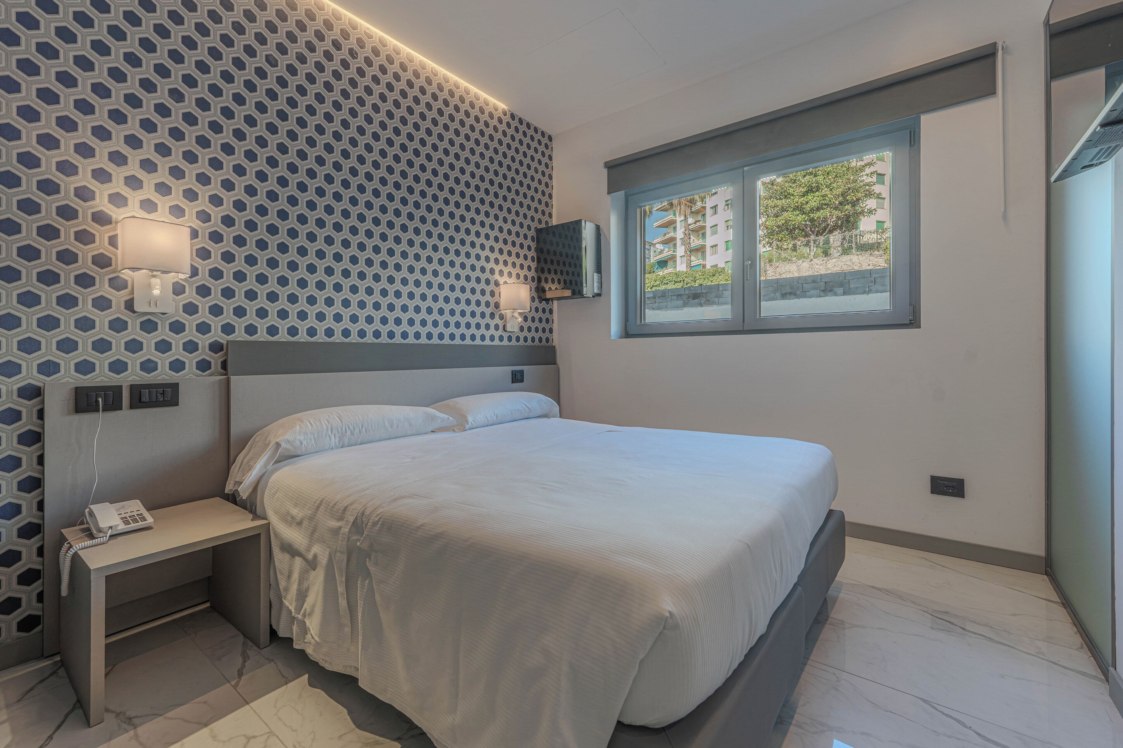 Quarto moderno com cama grande, mesa de cabeceira com telefone, televisão na parede e janela com vista para prédios.