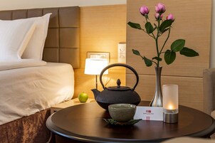 Quarto de hotel com uma cama, uma mesinha de cabeceira com um conjunto de chá, um vaso com flores e uma vela acesa.