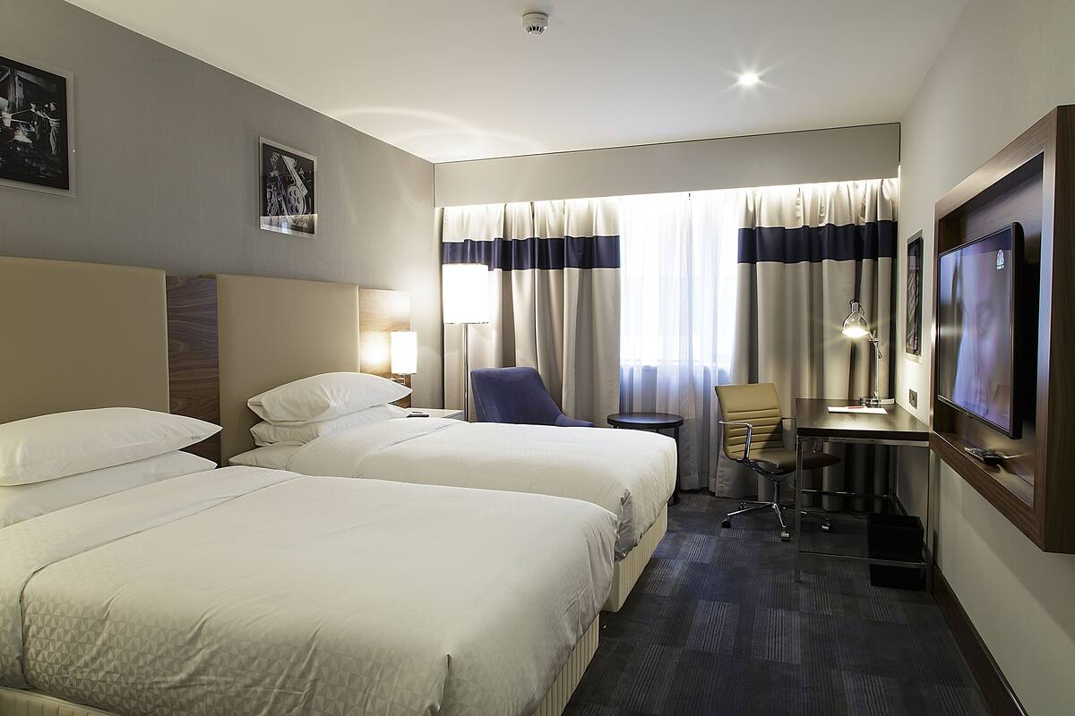 Quarto de hotel com duas camas, uma escrivaninha, uma cadeira, uma televisão e uma janela com cortinas.