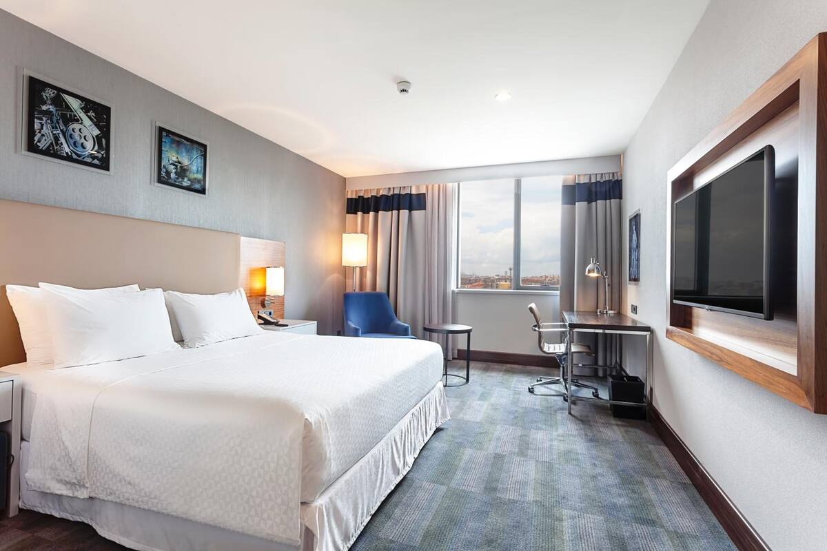 Quarto de hotel com uma cama grande, uma escrivaninha com cadeira, TV e vista para a cidade.