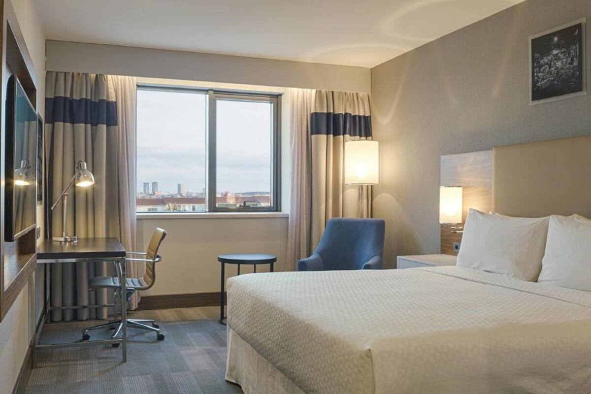 Quarto de hotel com cama, escrivaninha, cadeira, luminárias e vista para a cidade.