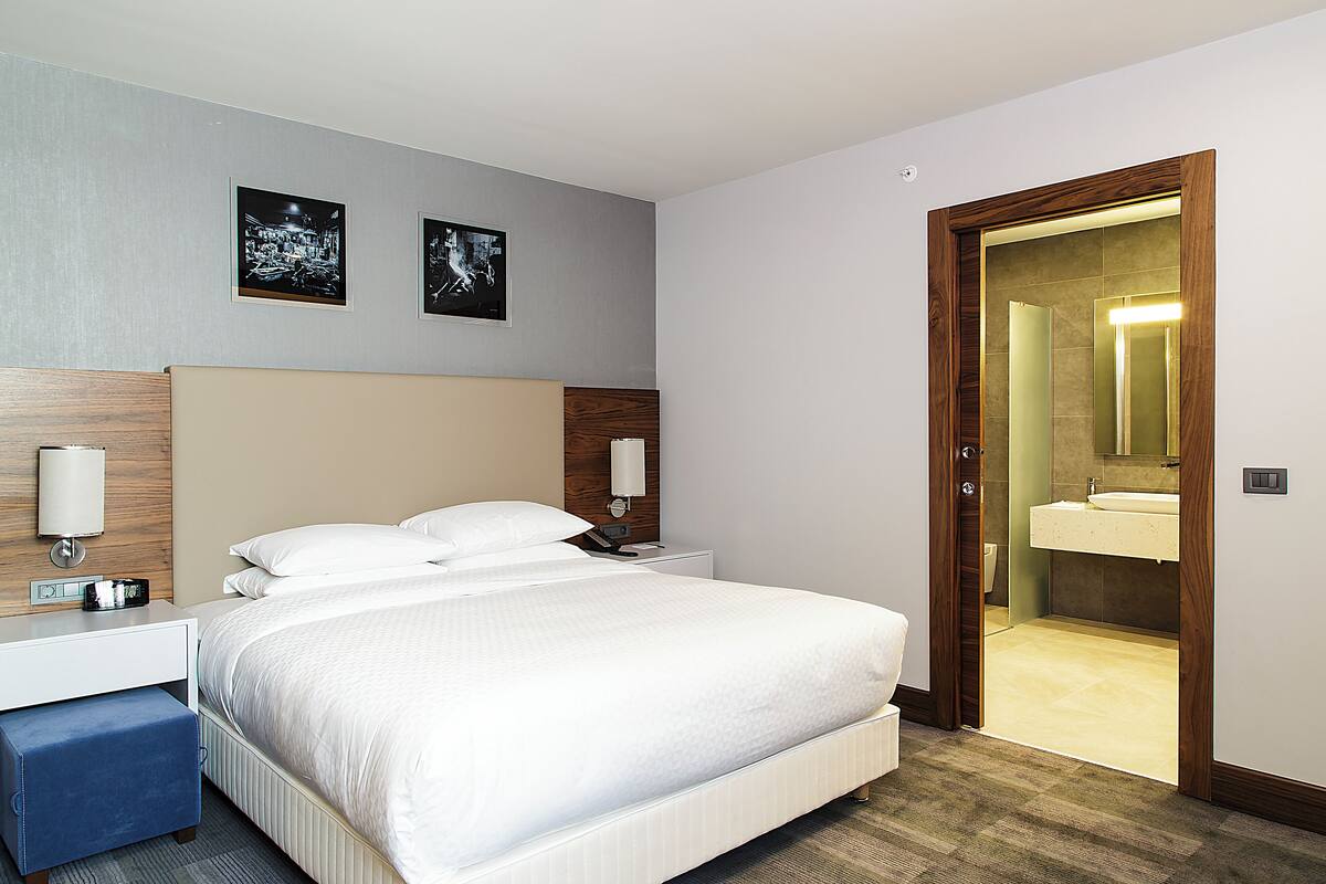 Quarto de hotel com uma cama grande, mesas de cabeceira, um banquinho e um banheiro visível através de uma porta aberta.