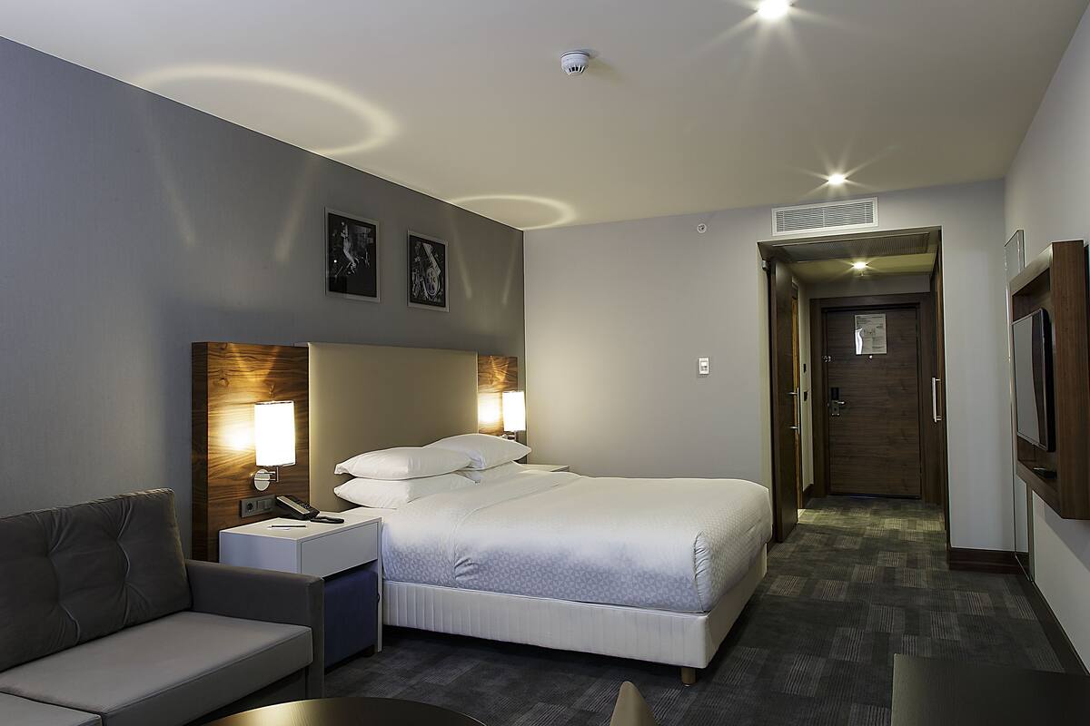 Quarto de hotel com uma cama, um sofá, uma escrivaninha com uma luminária, uma televisão e uma porta que dá para outro espaço.