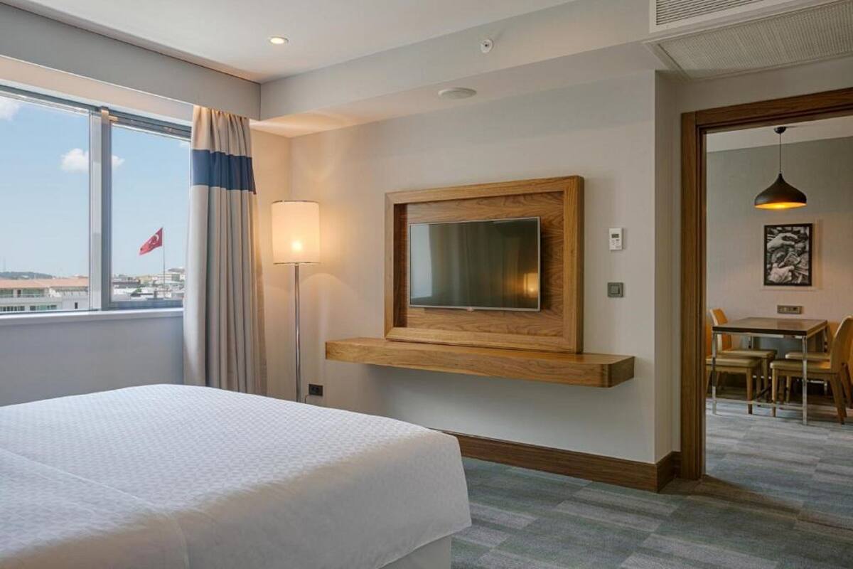 Quarto de hotel com uma cama, televisão na parede, luminária, área de refeições com cadeiras e uma mesa, e vista para a cidade através de uma janela.