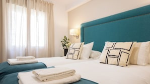Quarto de hotel com cama grande, cabeceira verde-água, roupa de cama branca e mesa de cabeceira com abajur e planta.