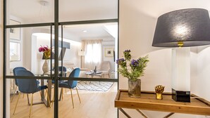 Uma sala de estar moderna com divisória de vidro, área de jantar com cadeiras azuis, mesa lateral de madeira com abajur e um vaso com flores.