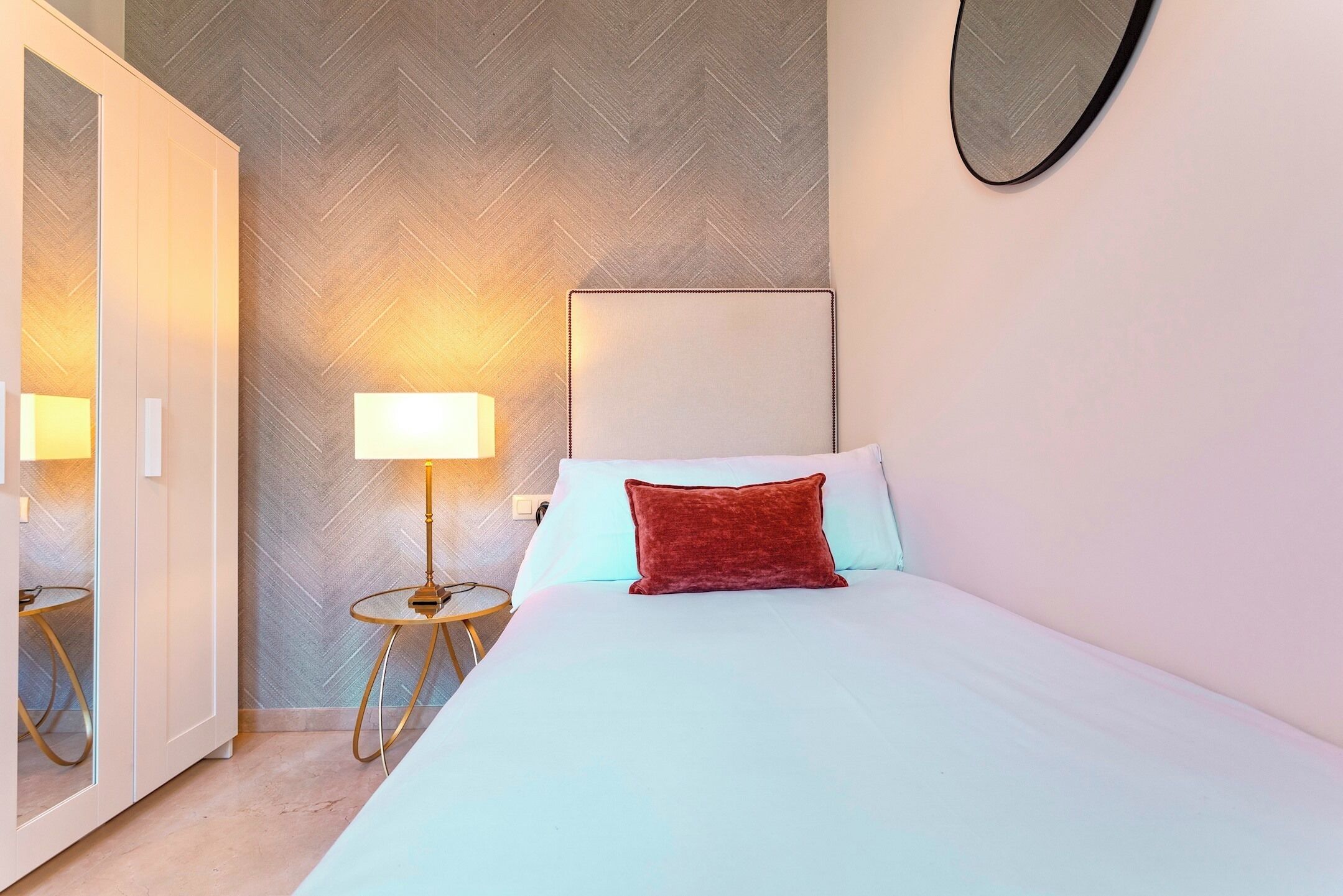 Quarto de hotel com cama, abajur, espelho e parede com padrão herringbone.