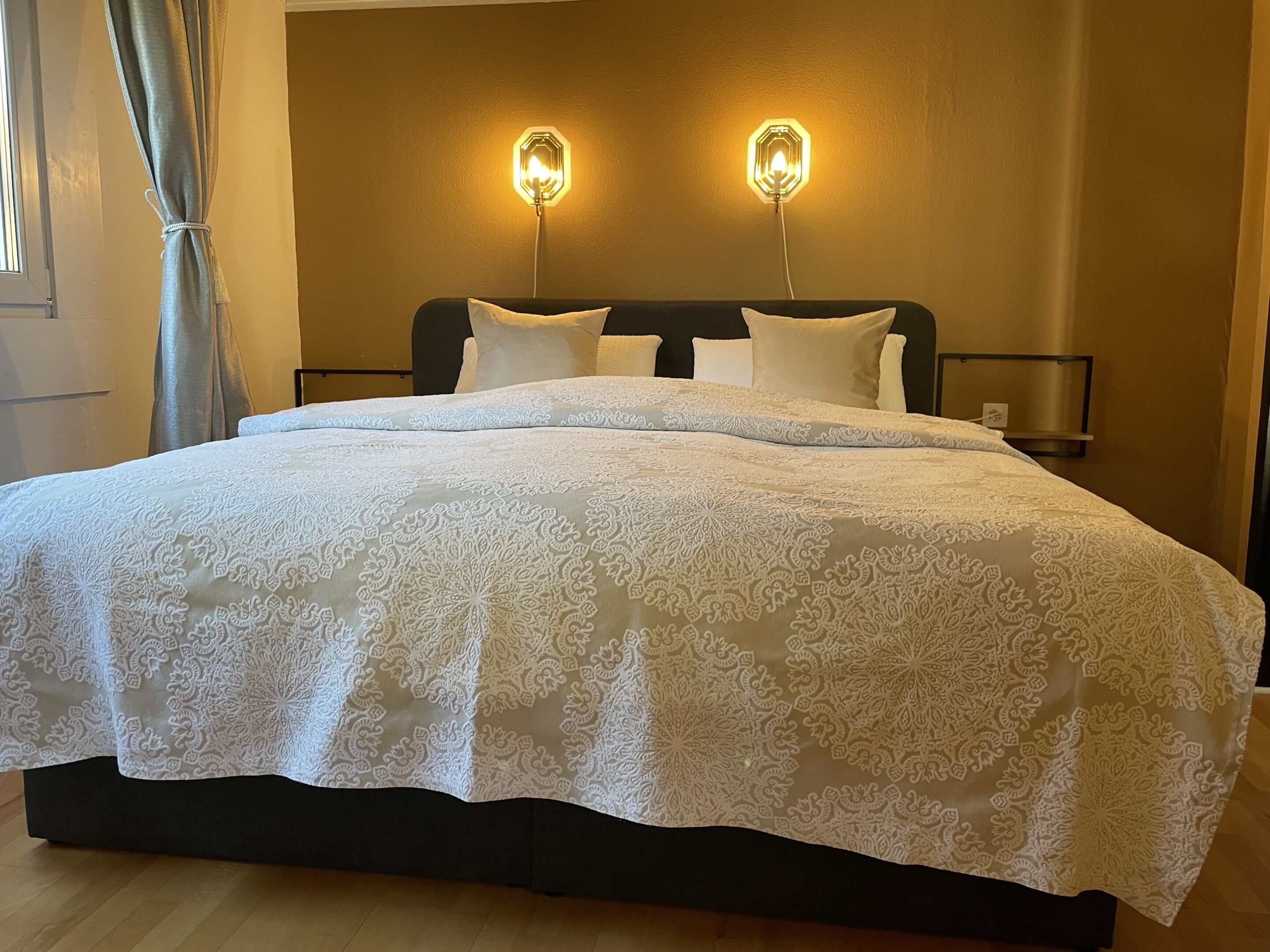 Cama bem arrumada com colcha estampada, duas luminárias fixadas na parede e mesa de cabeceira com um objeto em cima.